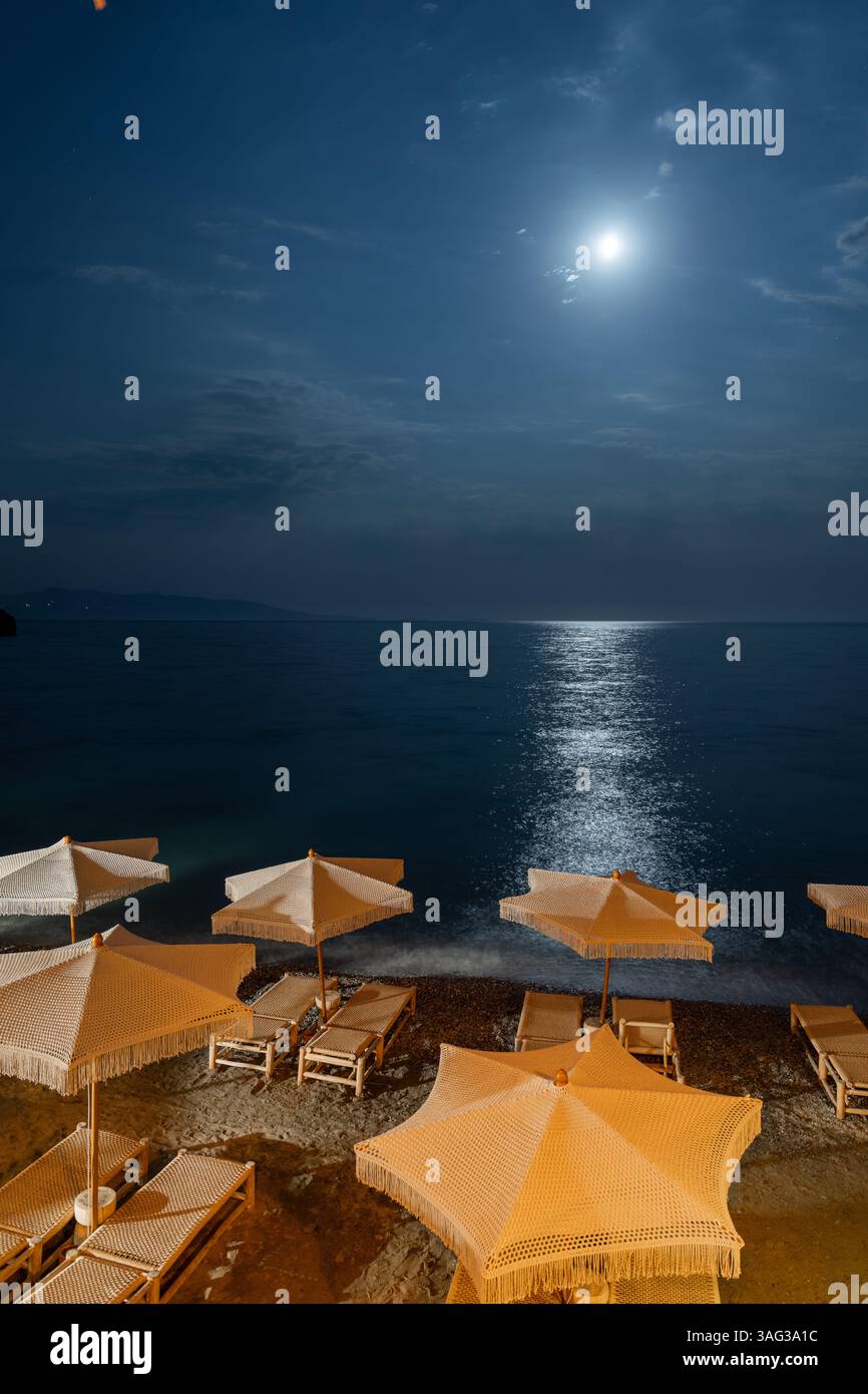 A peaceful moonlit beach with empty sun loungers and straw parasols ...