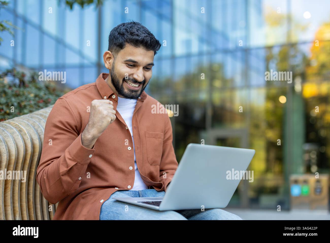 A smiling man sits outdoors with a laptop, clenching his fist in ...