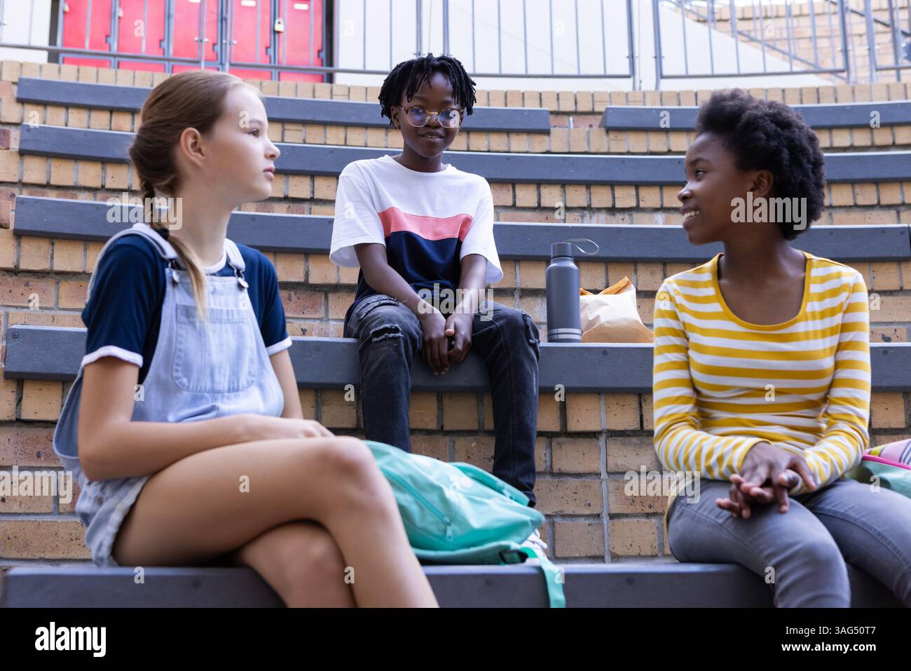 In school, three diverse kids sitting on steps, talking and relaxing ...