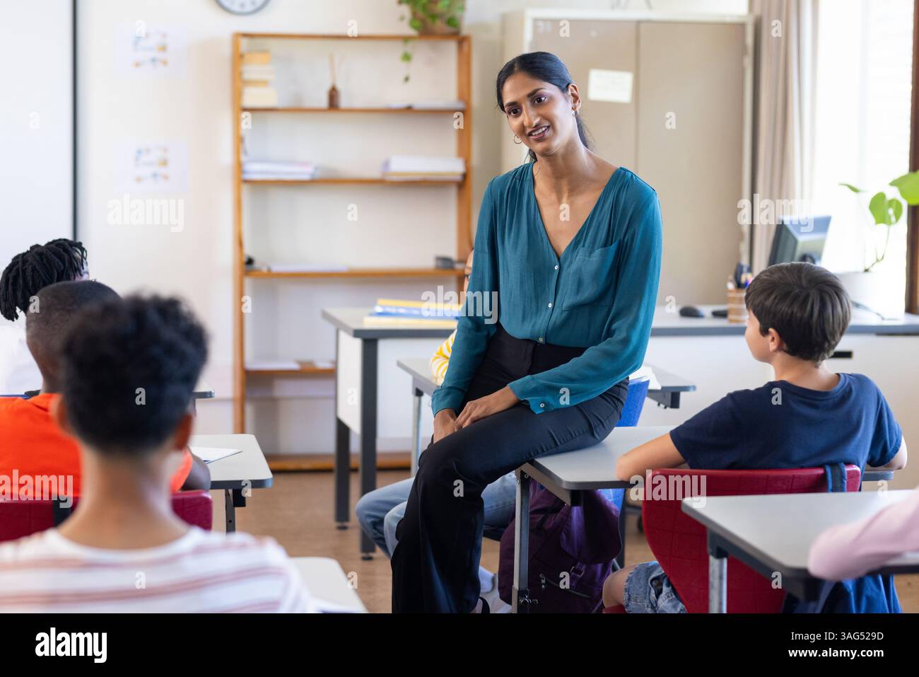 In school, Indian female teacher sitting on desk and engaging with ...