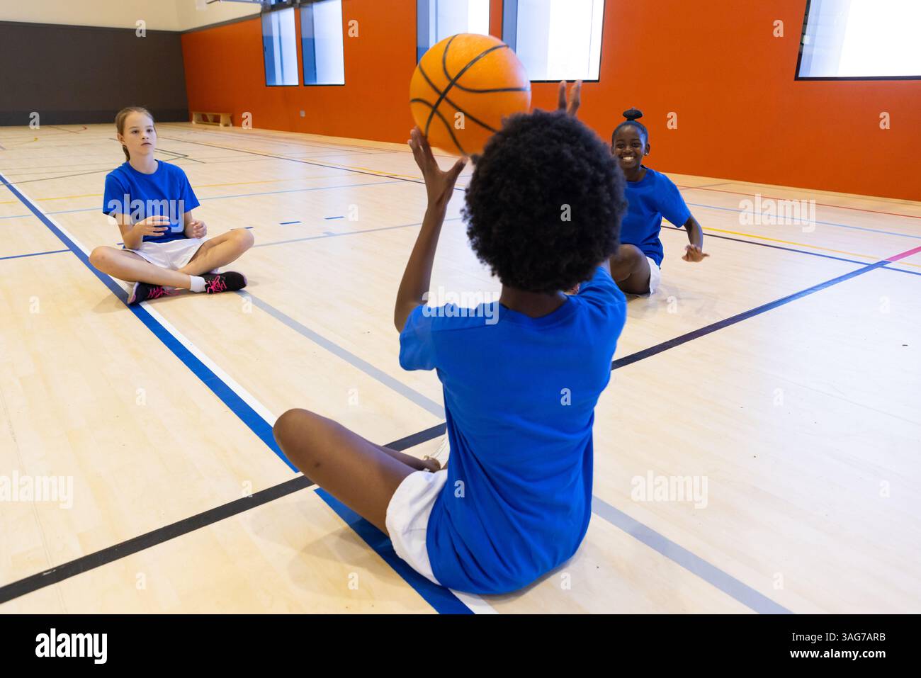 Playing basketball, three diverse girls sitting on gym floor in school ...