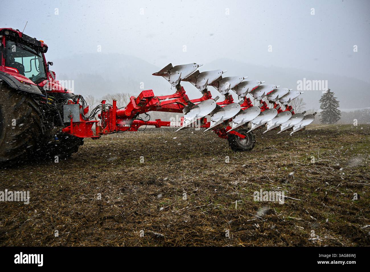 Red tractor pulling plow in field during snowfall, preparing soil for ...