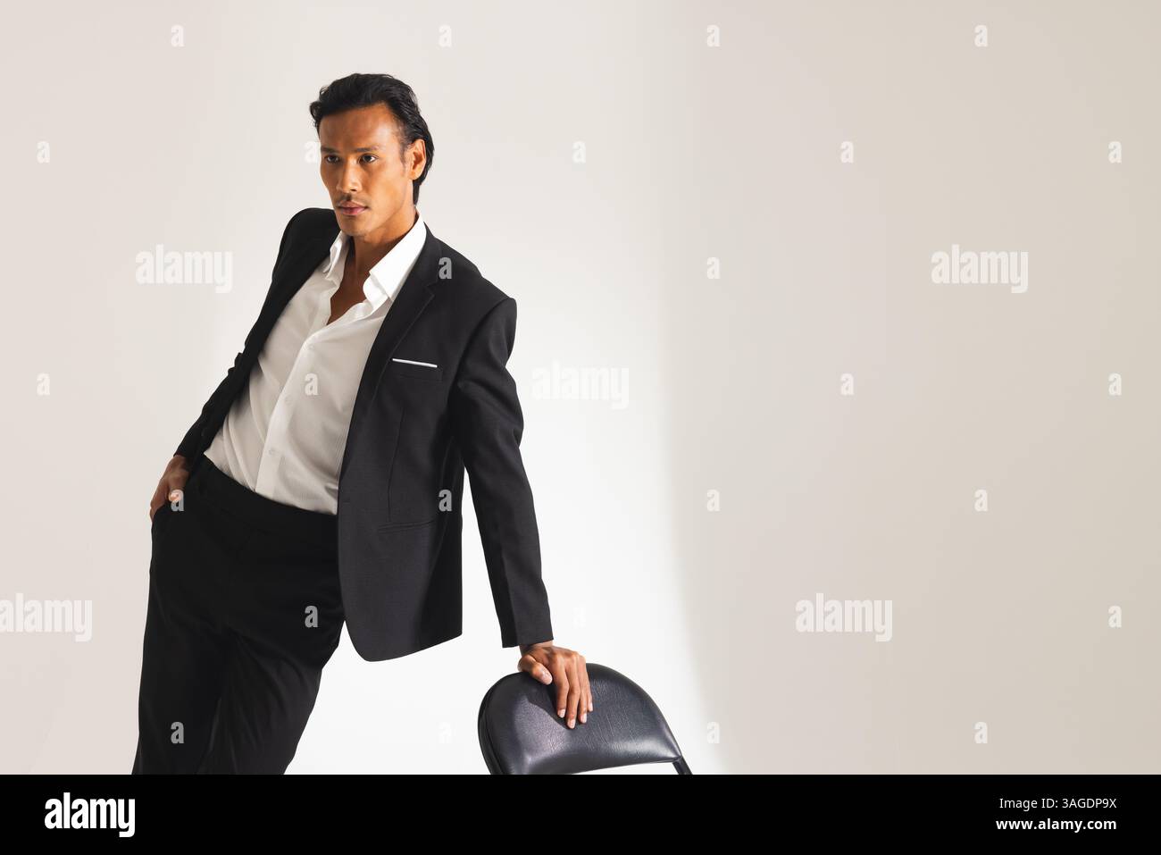 Confident man in black suit leaning on chair, posing in fashion studio ...