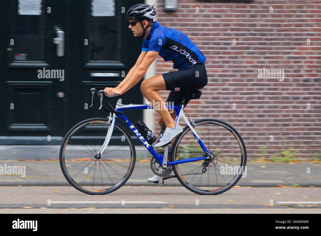 A cyclist riding a blue and white Trek road bicycle, wearing a "BJÖRKA ...