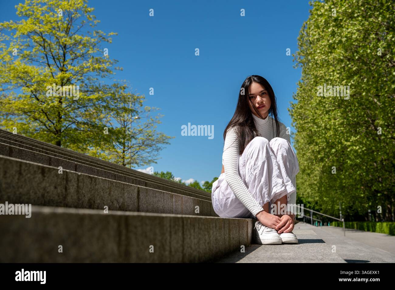 Gen Z woman in a white outfit is sitting on outdoor concrete stairs and ...