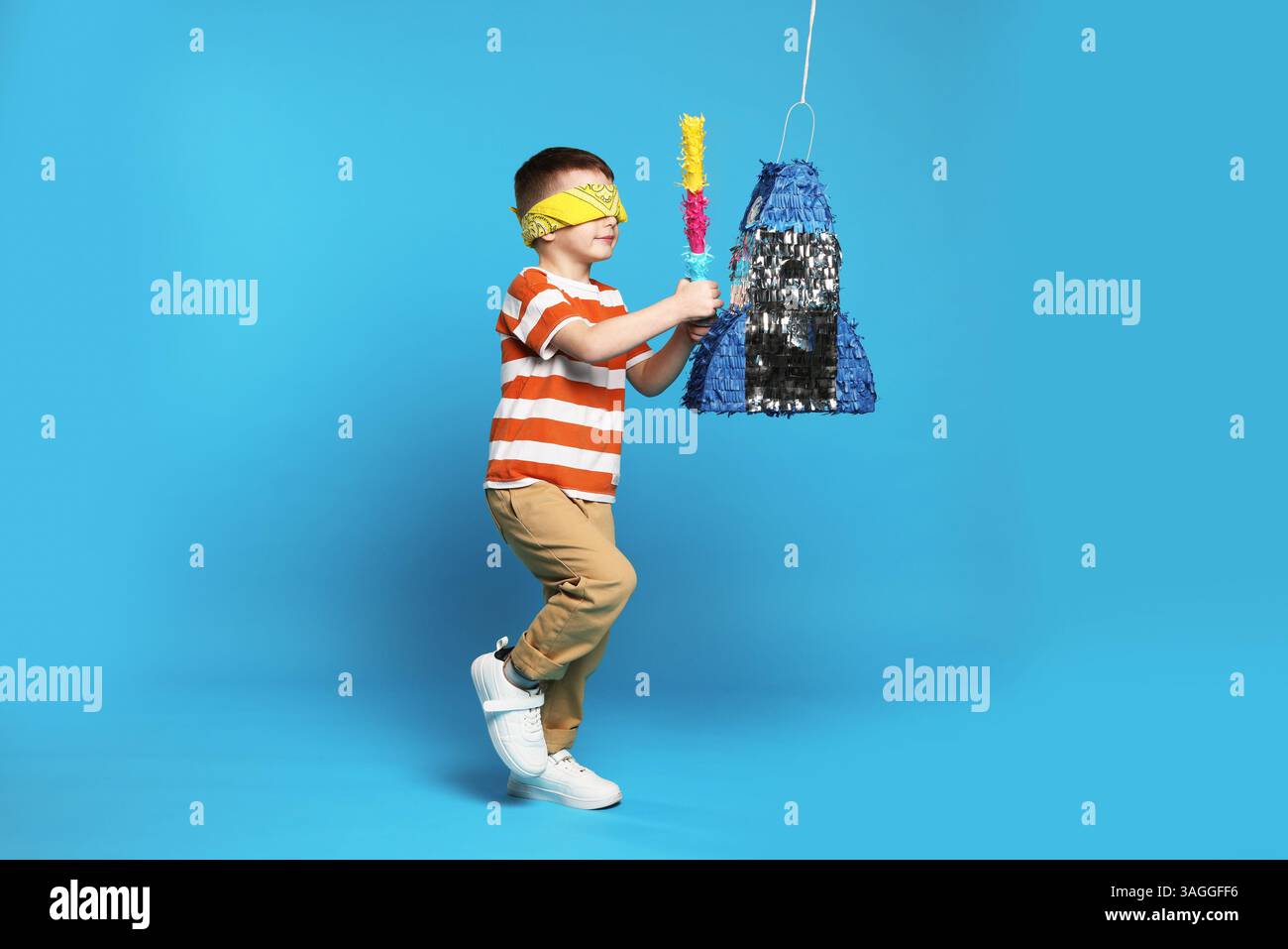 Cute boy with tied eyes breaking rocket shaped pinata on light blue ...