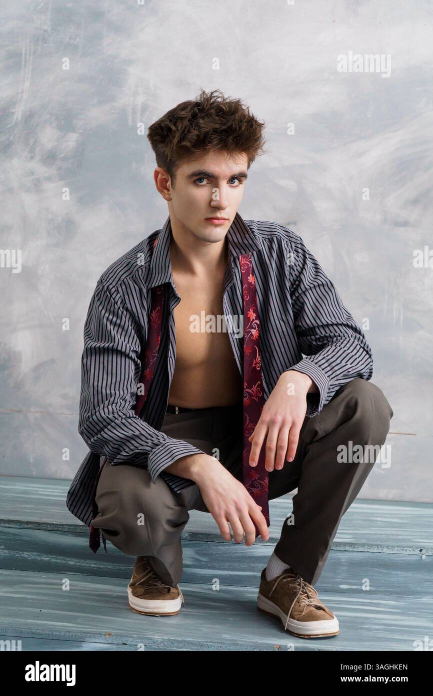 Young man in unbuttoned shirt and tie crouching against textured grey ...