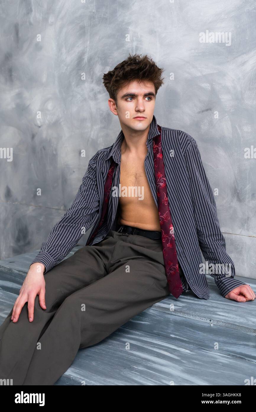 Young man with unbuttoned shirt and red tie posing seated on gray ...