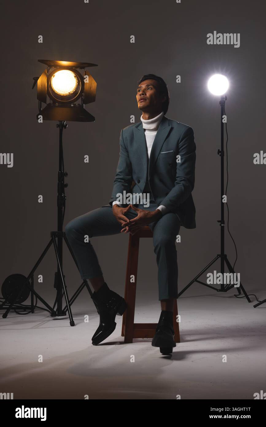 Fashion model in stylish suit sitting on stool under studio lights ...