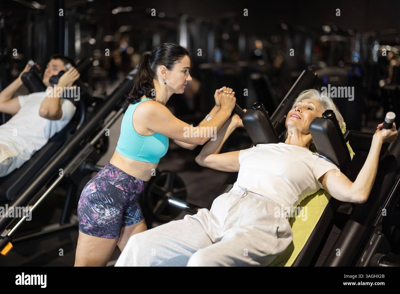 Senior woman performing squats on hack machine with coach Stock Photo ...