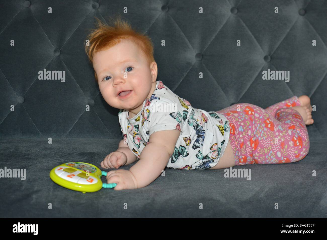 Red hair baby girl lying on a couch on a belly and holding a toy. 6 ...