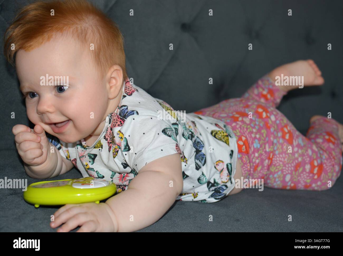 Red hair baby girl lying on a couch on a belly and holding a toy. 6 ...