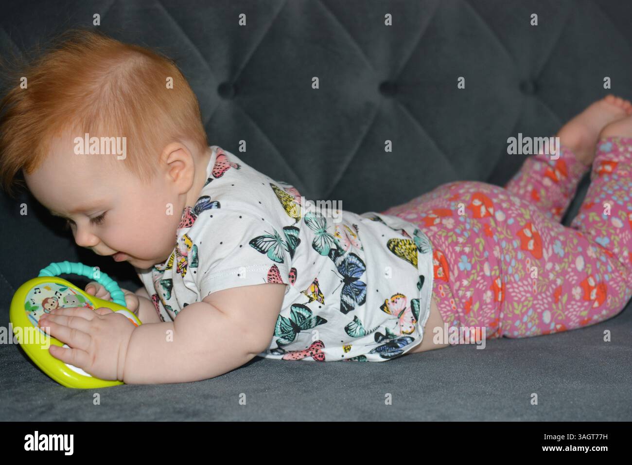 Red hair baby girl lying on a couch on a belly and holding a toy. 6 ...
