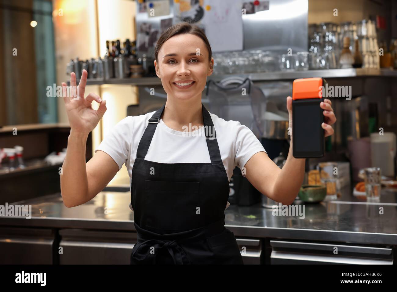 Smiling cafe worker with payment terminal showing OK gesture indoors ...
