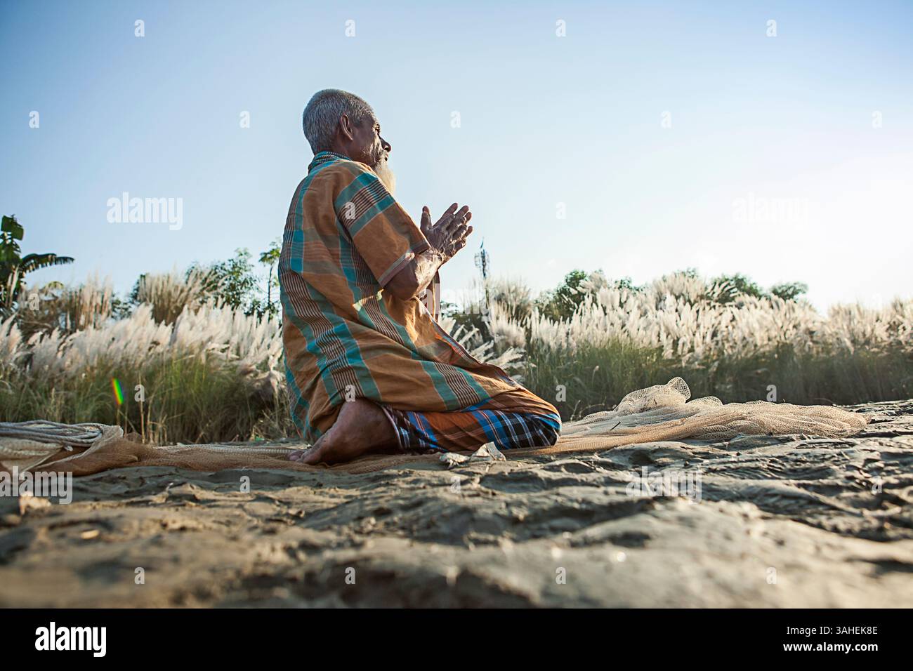 An elderly Bangladeshi man prays outdoors at dawn, seated on a ...