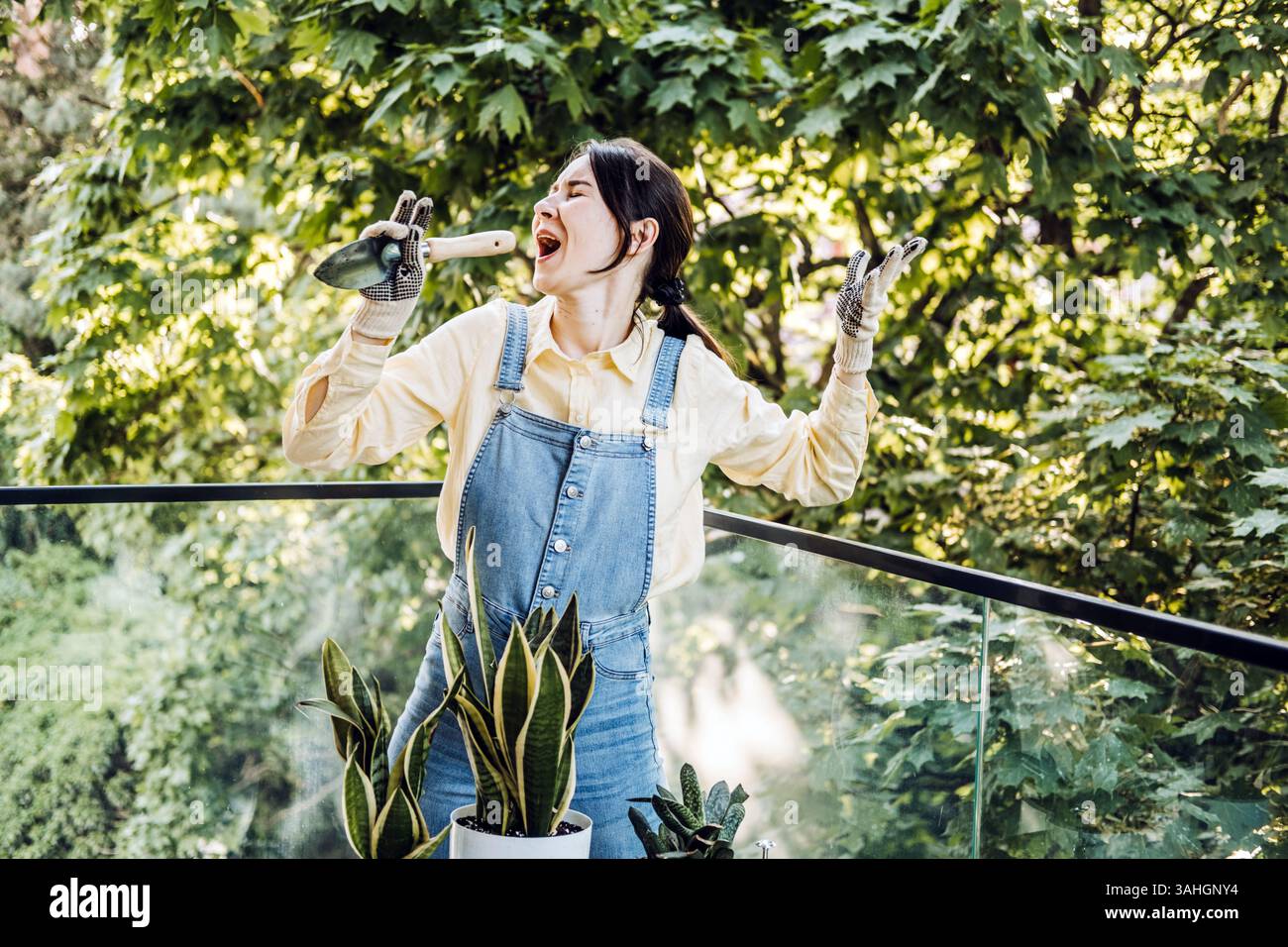 Young Asian woman playfully singing into a garden trowel while caring ...