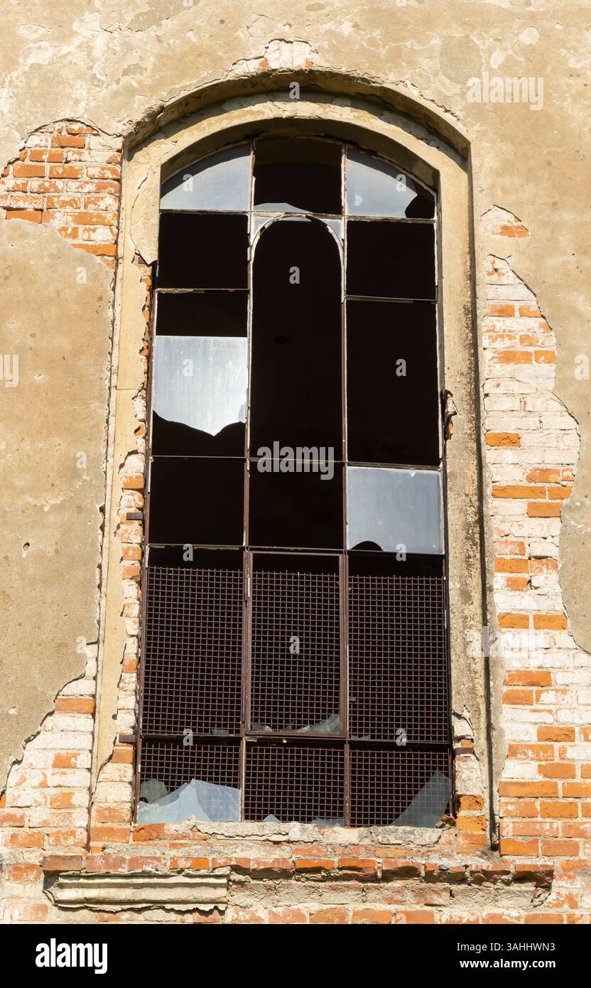 A tall, arched window with broken glass panes is set in a weathered ...