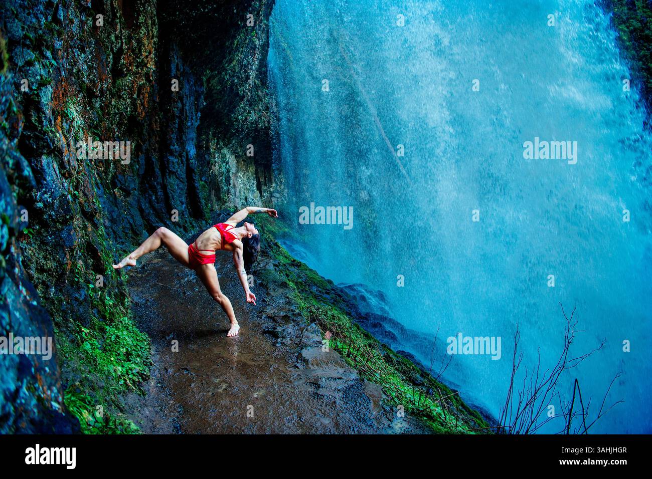 Dancer in red poses gracefully by a stunning waterfall on a rocky and ...