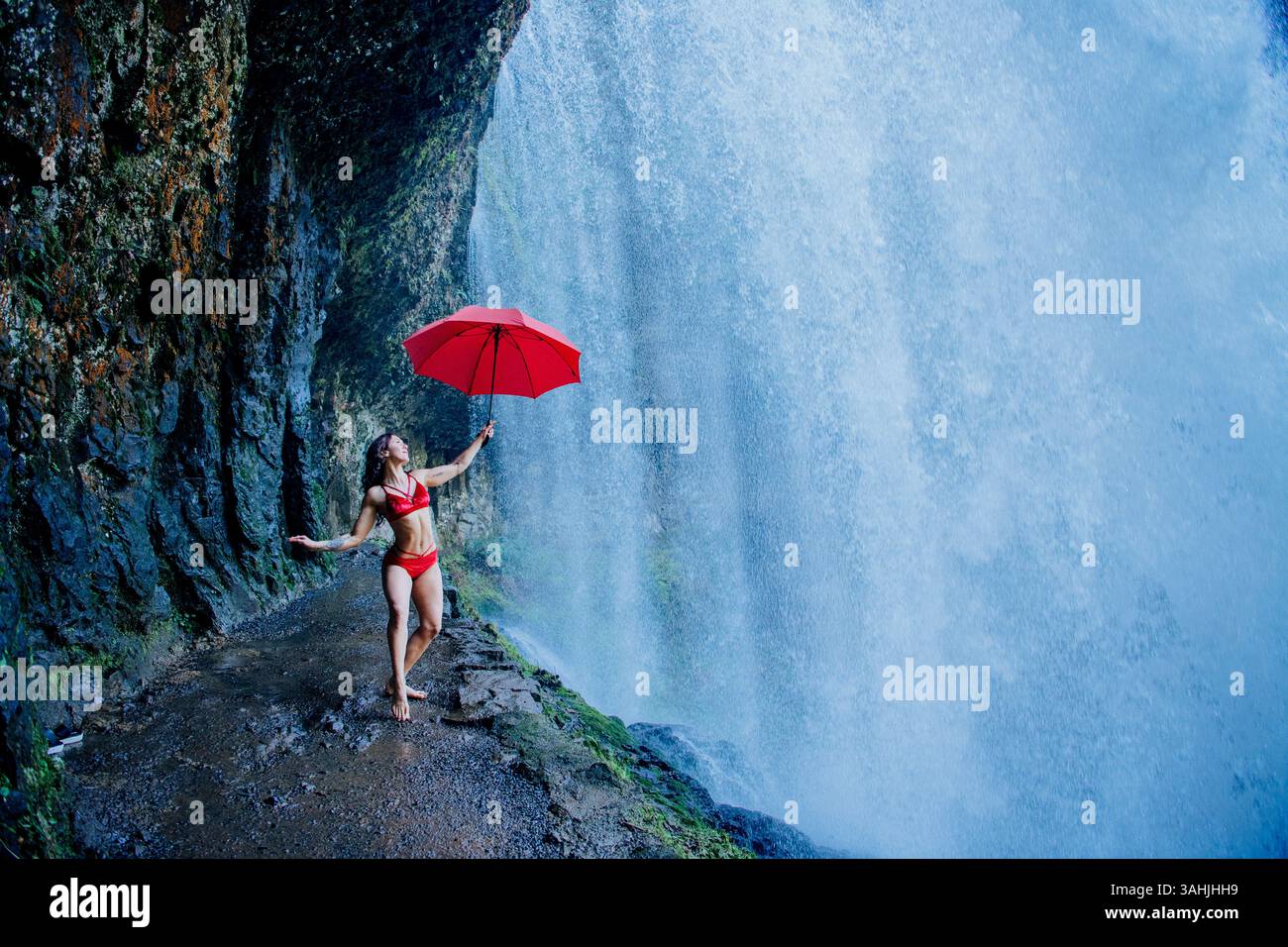 Woman in red bikini holding umbrella beneath a waterfall on rocky path ...