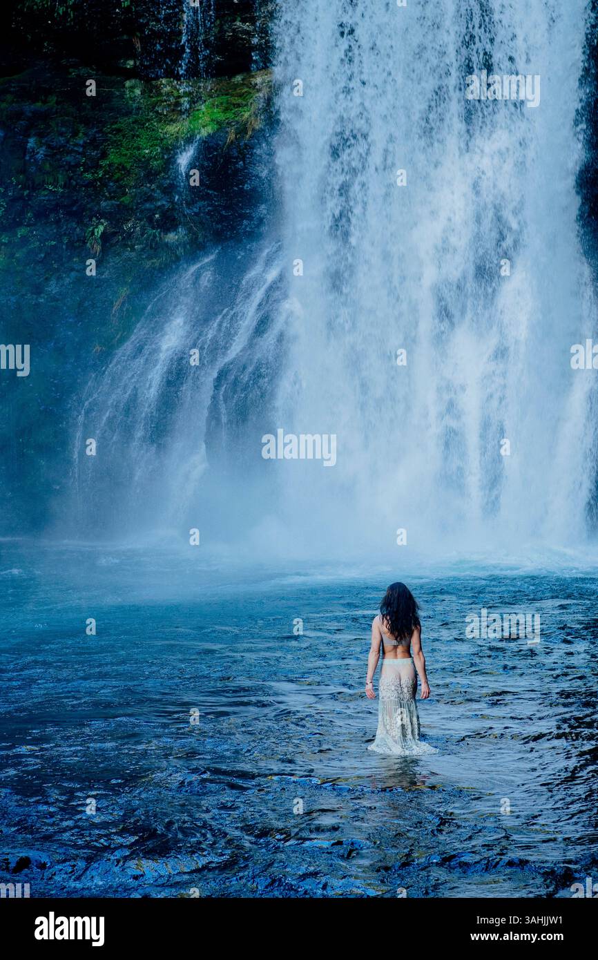 A woman in a white dress stands in front of a majestic waterfall ...