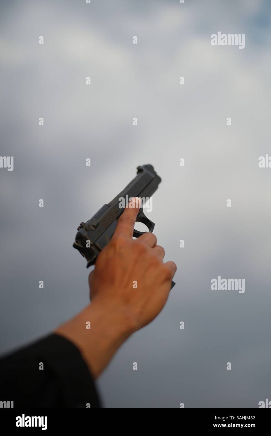 A hand holding a pistol pointed skyward against a cloudy sky backdrop ...