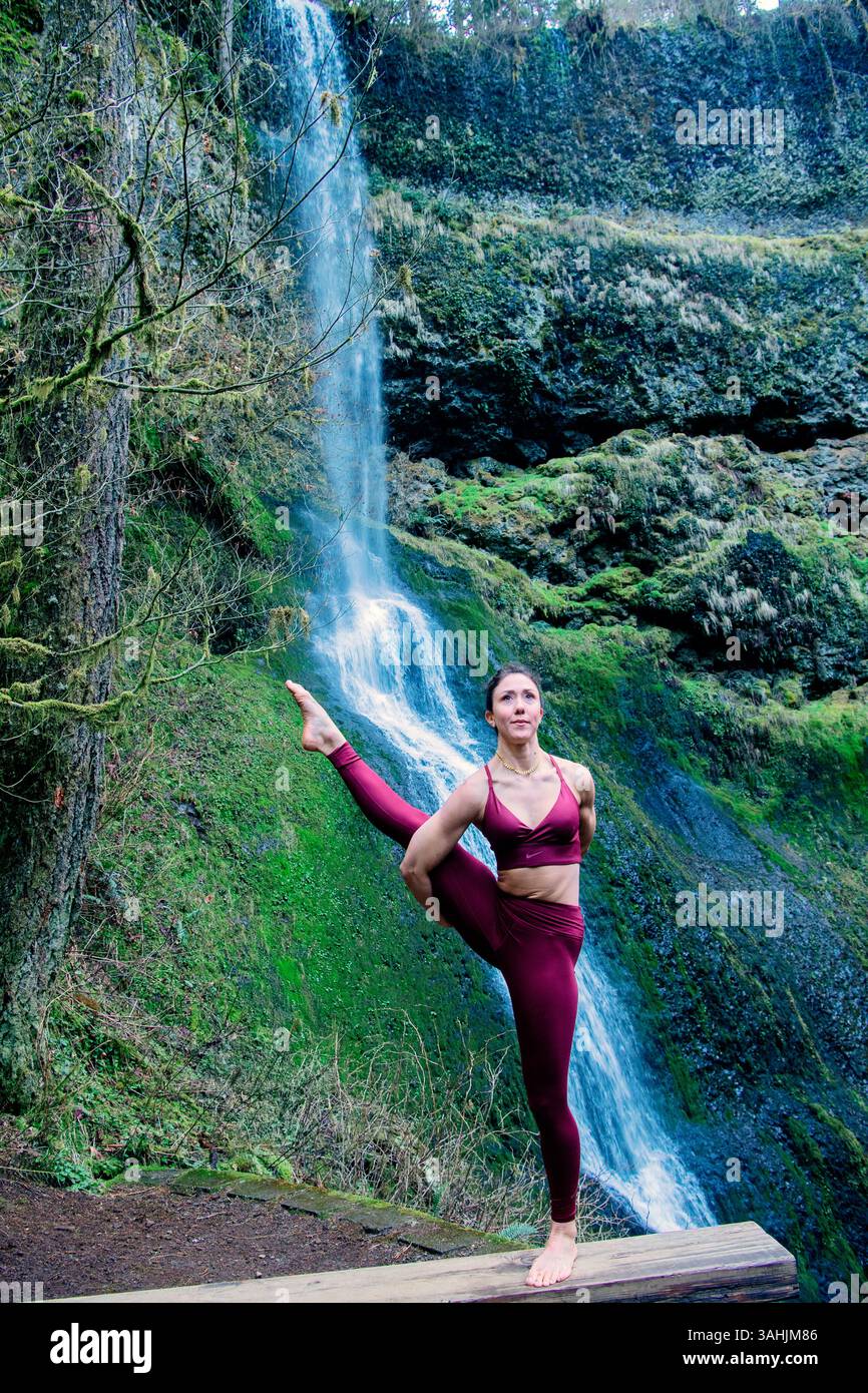 Woman practicing yoga in nature near a tall waterfall cascading down a ...