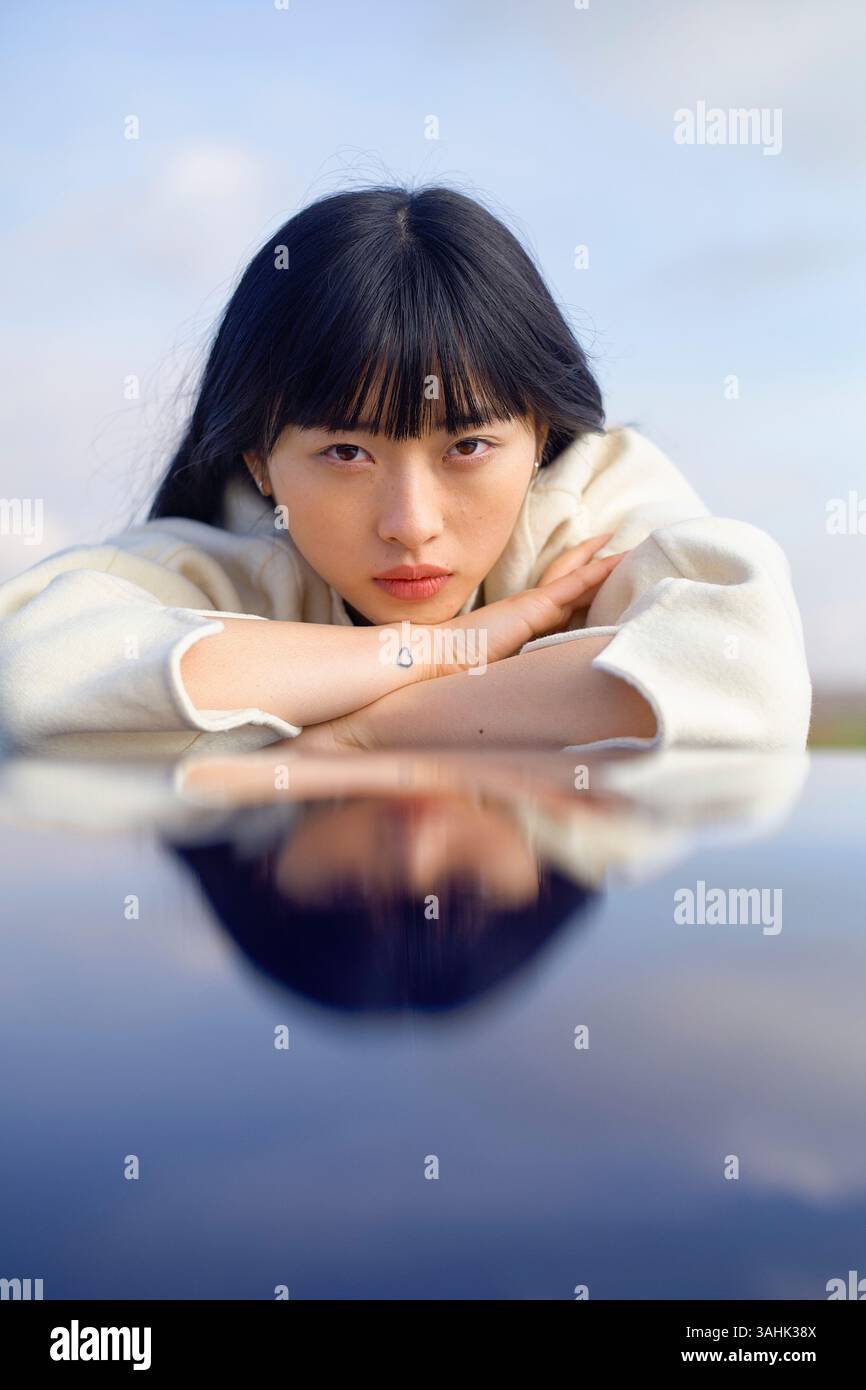Woman with black hair leaning on a reflective surface, with a distant ...