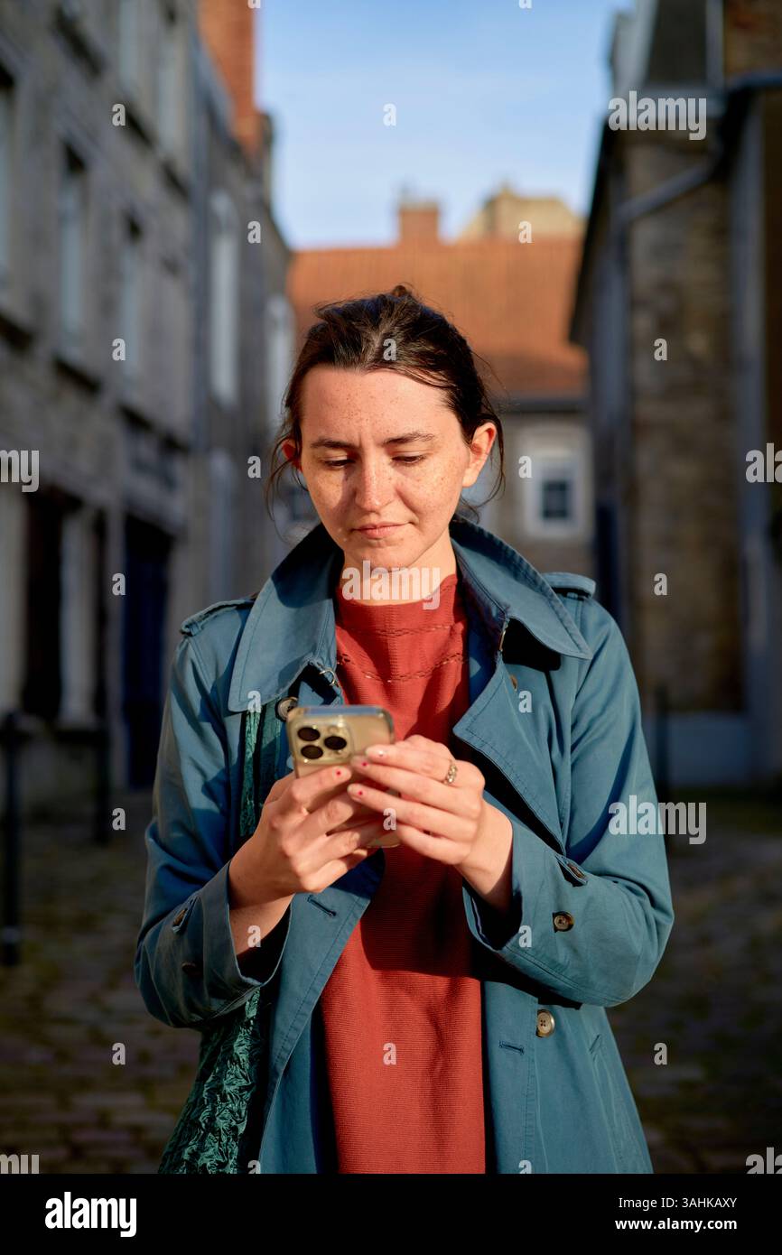 Woman in blue coat using smartphone on a cobblestone street with old buildings. Normandy, France ...