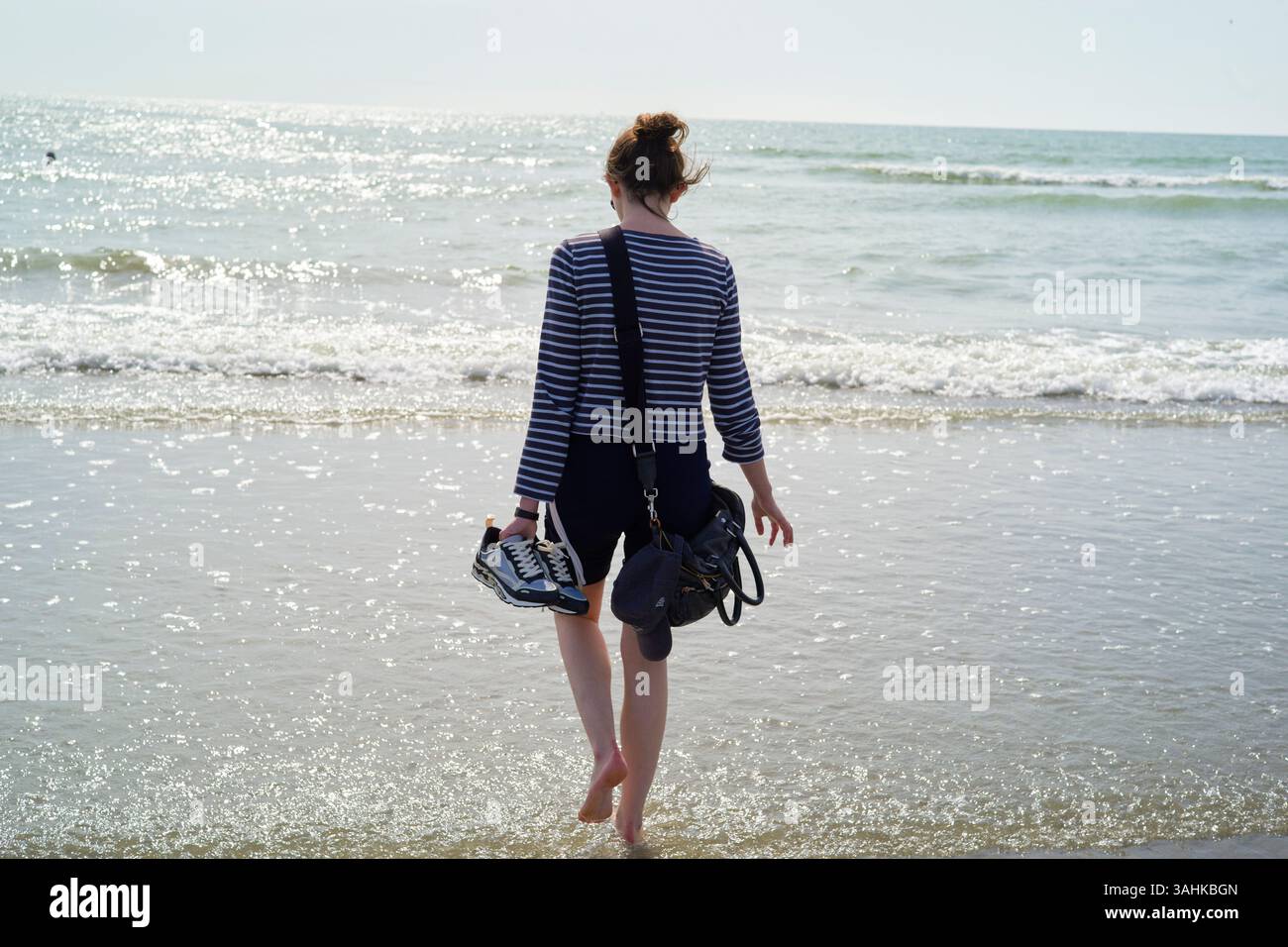 Woman in striped shirt walks barefoot on beach, carrying shoes and bag ...