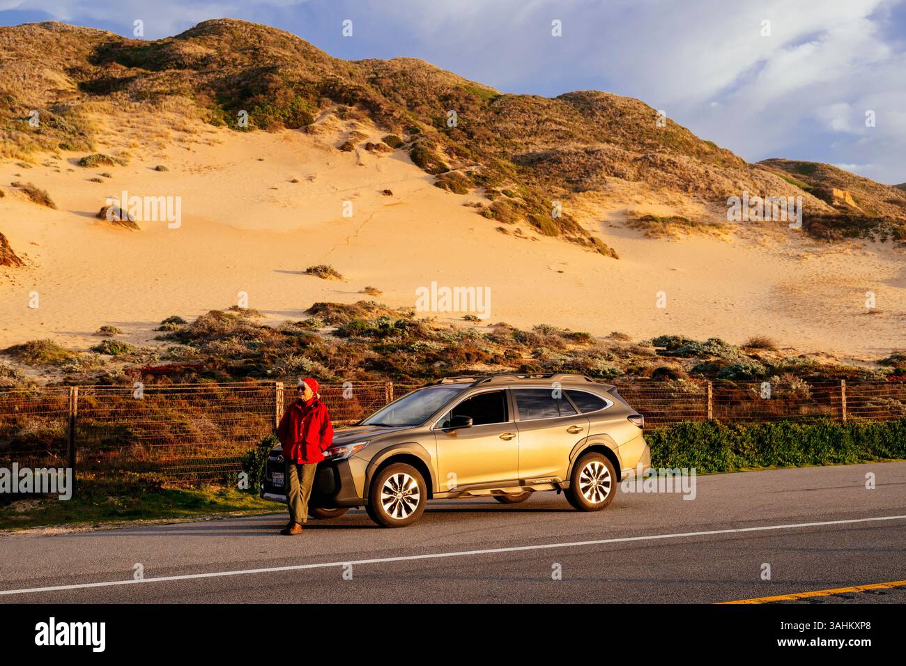 A woman stands by a golden SUV parked on a scenic desert road with sand ...