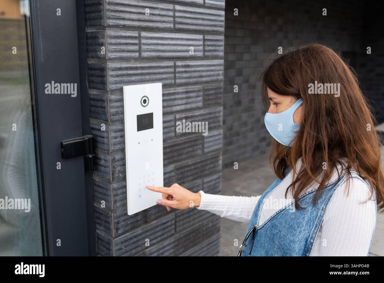 A young woman wearing a face mask uses a digital door intercom outside ...