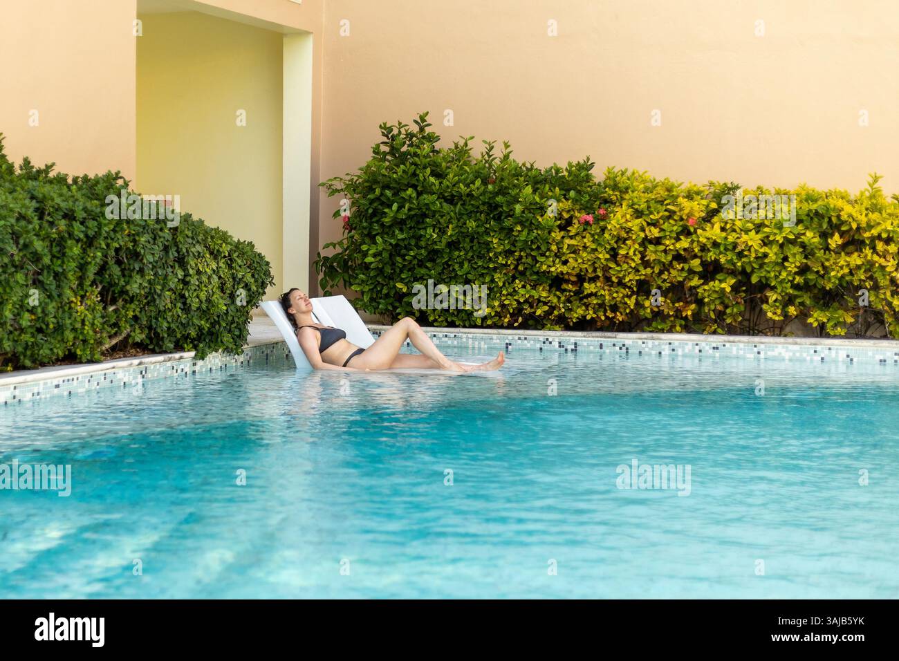 A young woman enjoys a quiet moment lounging by a bright blue pool, soaking up the sun in a lush ...