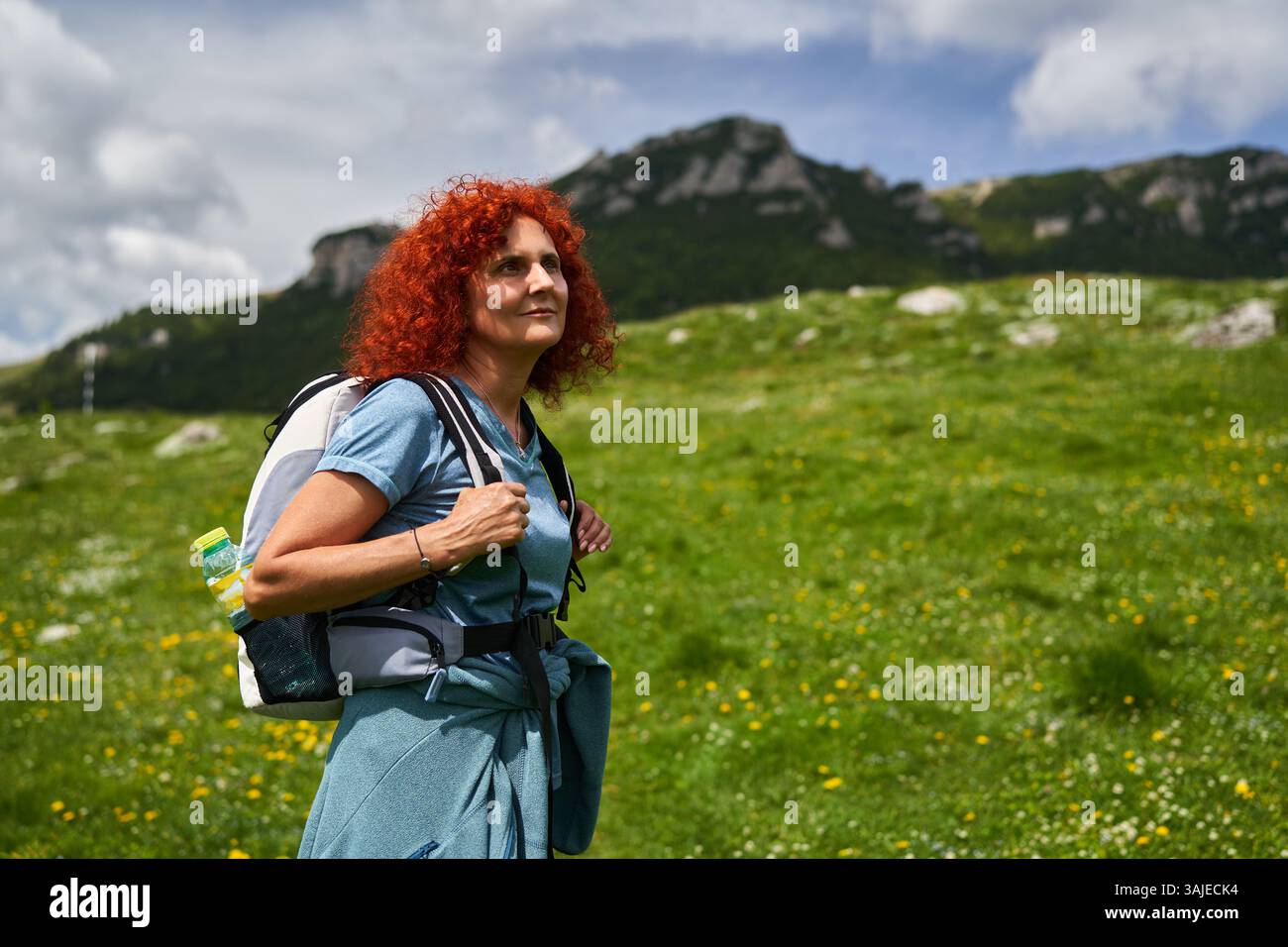 Middle aged redhead woman hiking in a green mountain meadow with backpack under a cloudy sky ...