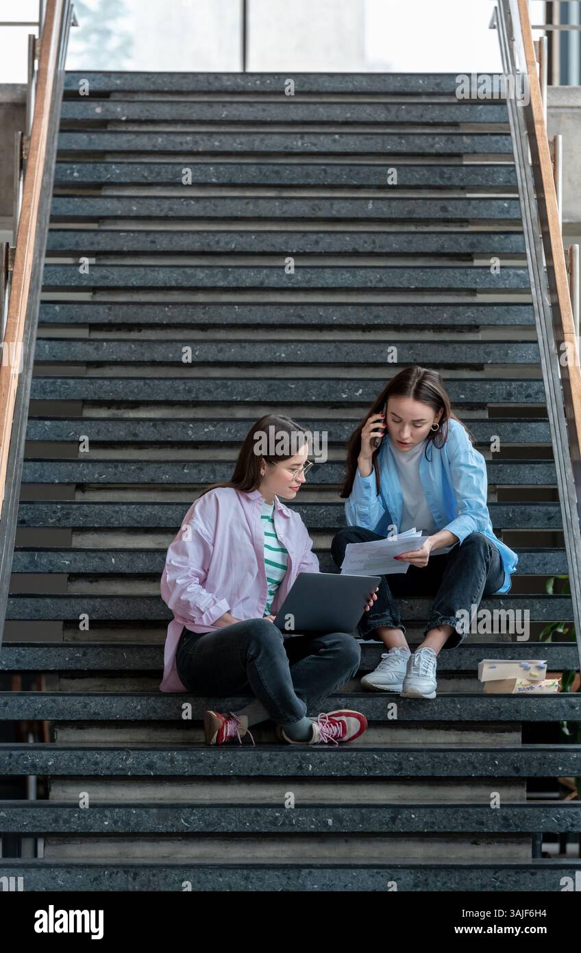 Two female students work side by side while sitting on the stairs in ...