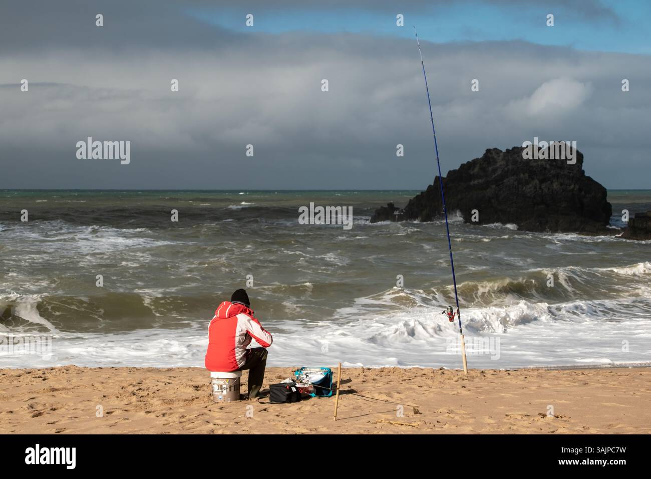 Beach fishing scene with male angler sitting on sand focused on ...