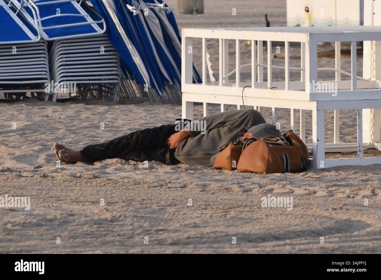 Rough sleeper on sandy beach in Miami, sea behind him, warm coastal ...