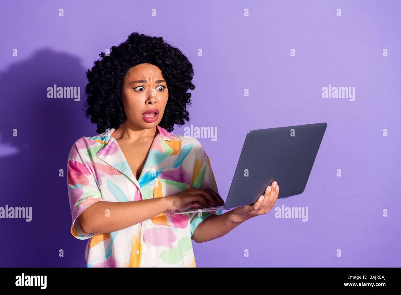 Surprised young woman holding laptop against violet background, wearing ...