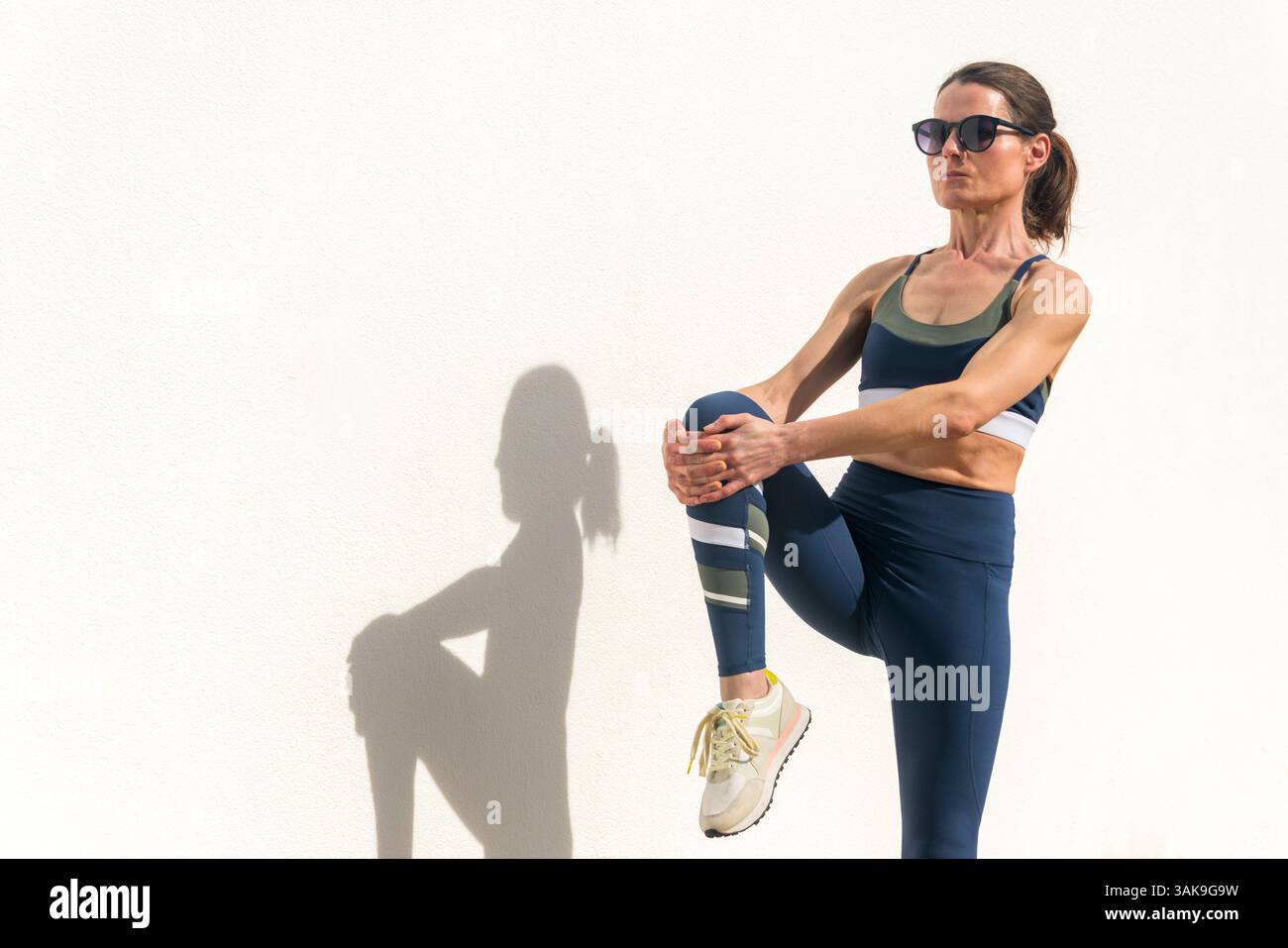 Sporty woman doing a leg stretch warm up exercise against a white wall ...