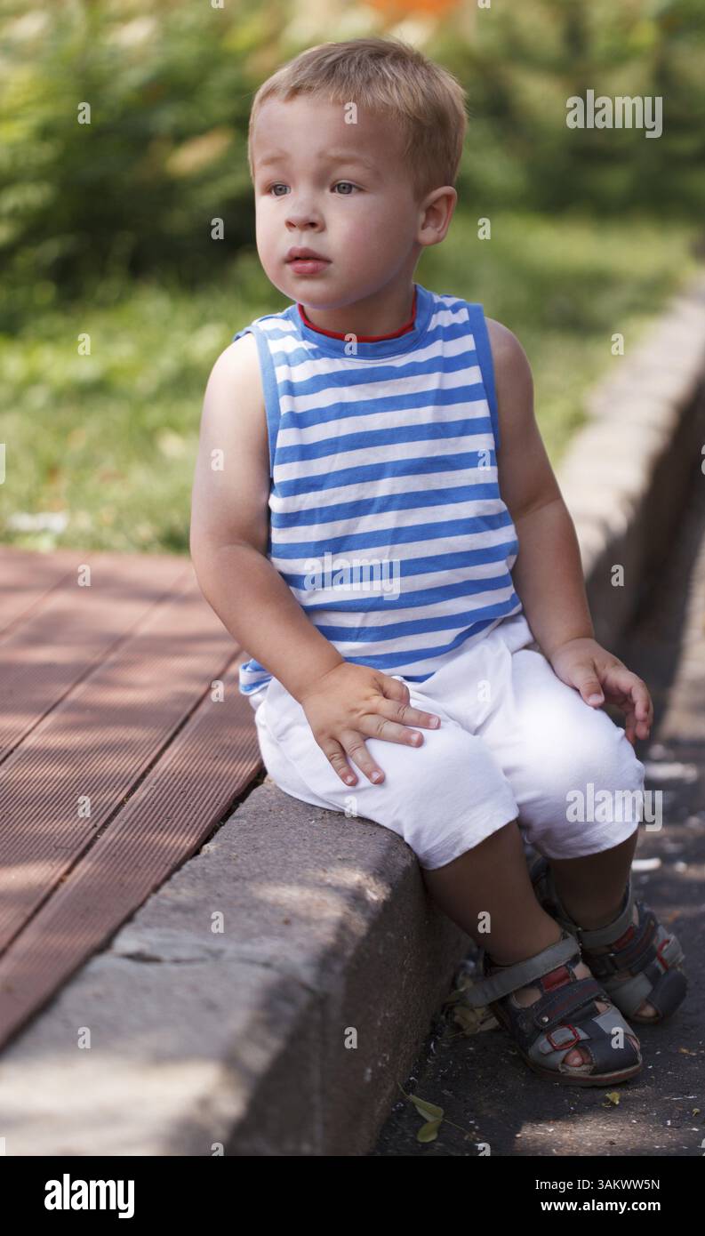 Portrait of a three year-old boy sitting on the curb and looking left ...