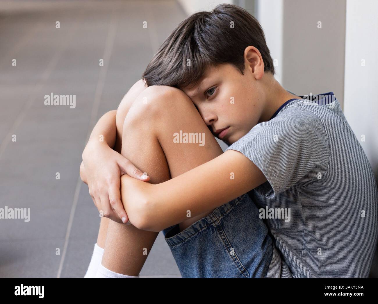 Sitting alone, boy hugging knees and looking sad in school hallway ...
