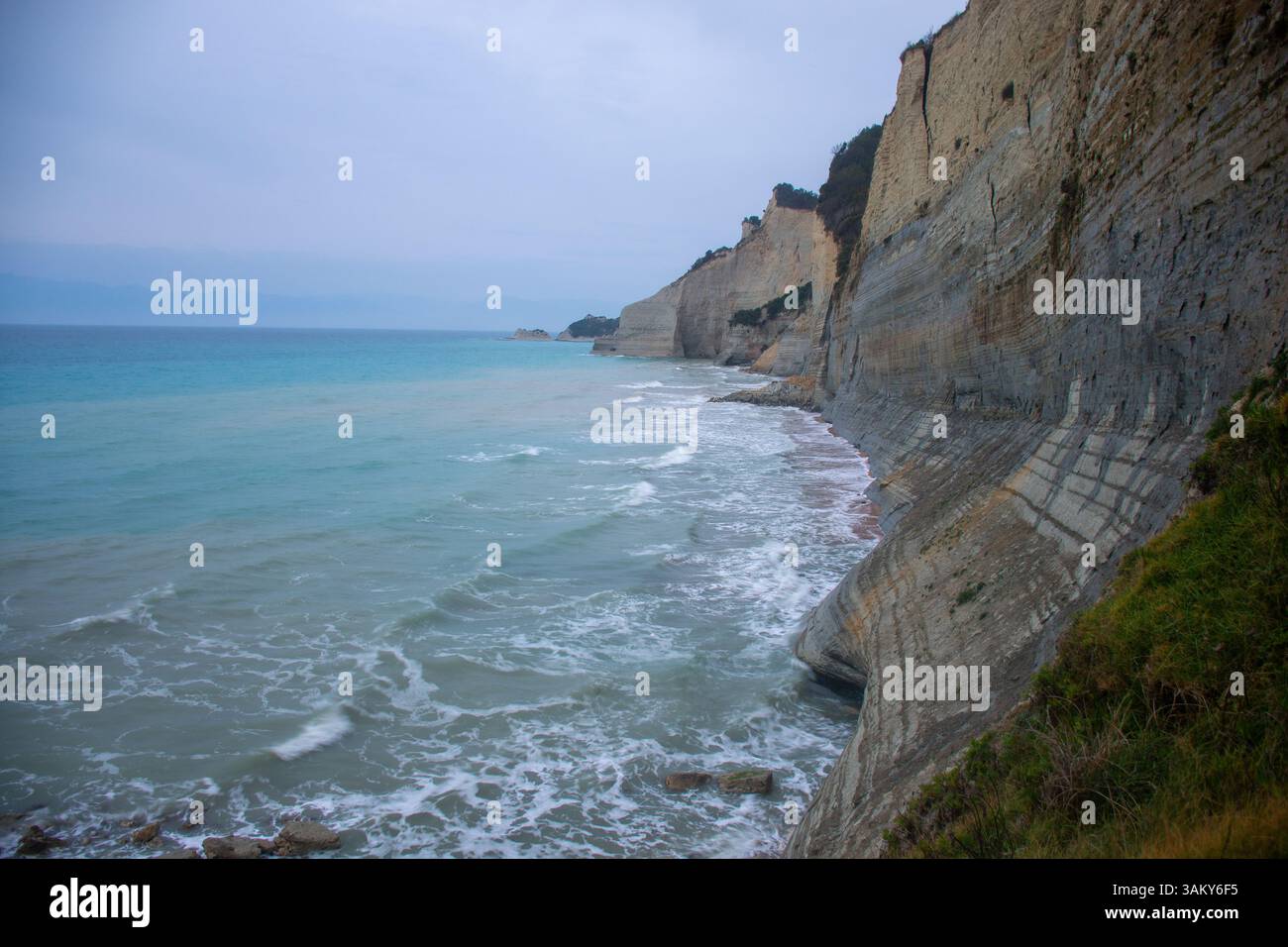 The towering cliffs meet the turquoise sea under a cloudy sky creating ...
