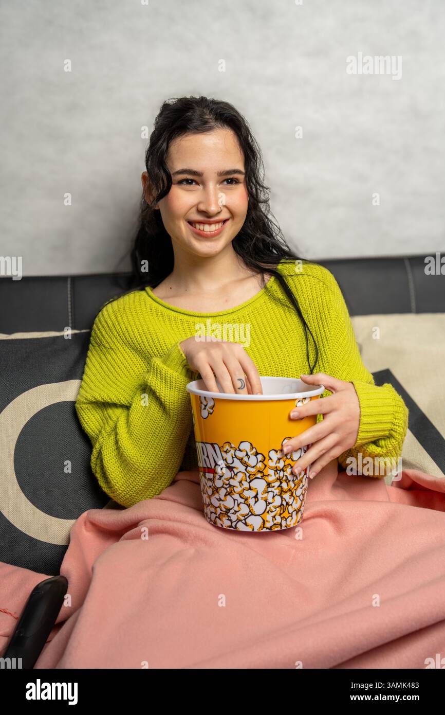 Smiling young woman enjoying a movie night at home, eating popcorn from a big bucket Stock Photo ...