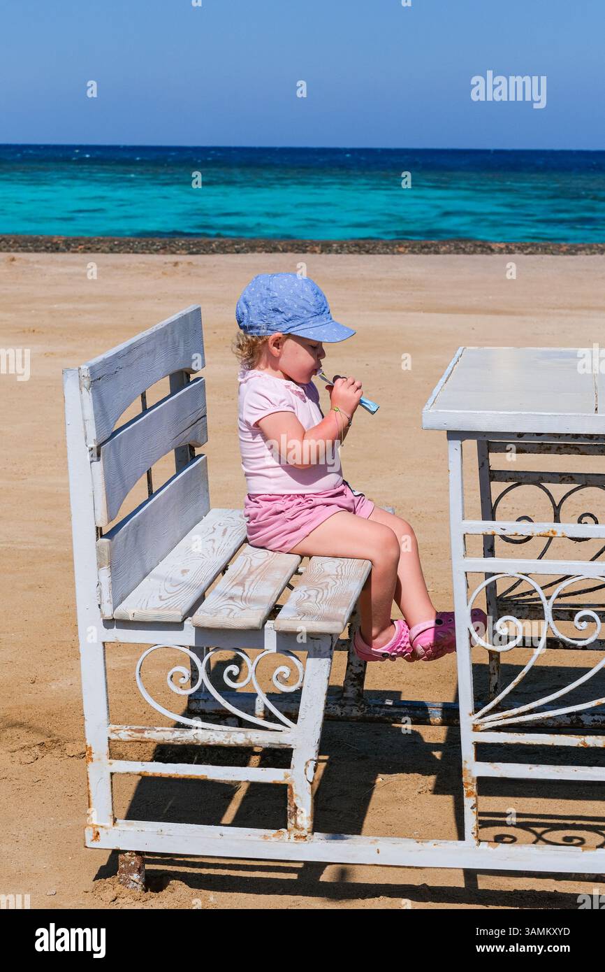 A little girl is sitting on a bench next to a table. She is brushing ...