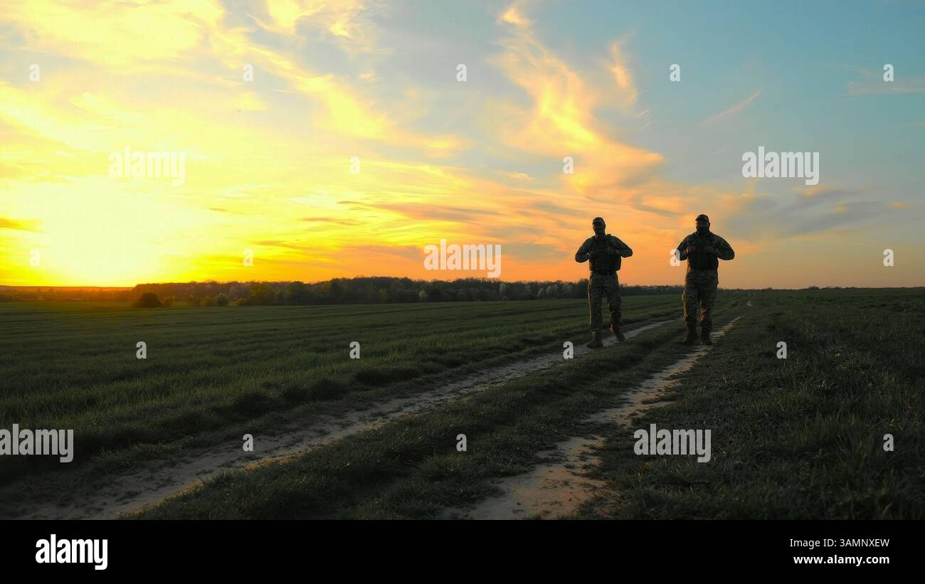 Soldiers walking along a dirt path during sunset. Two soldiers in full military attire walk ...
