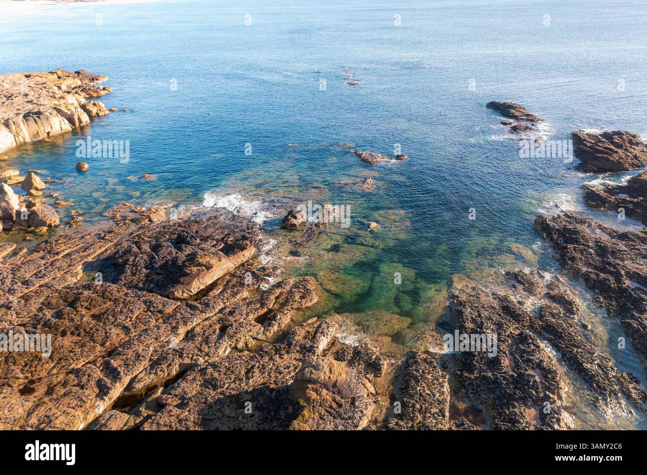 Aerial view of rocky shoreline and clear blue sea with surfers and ...