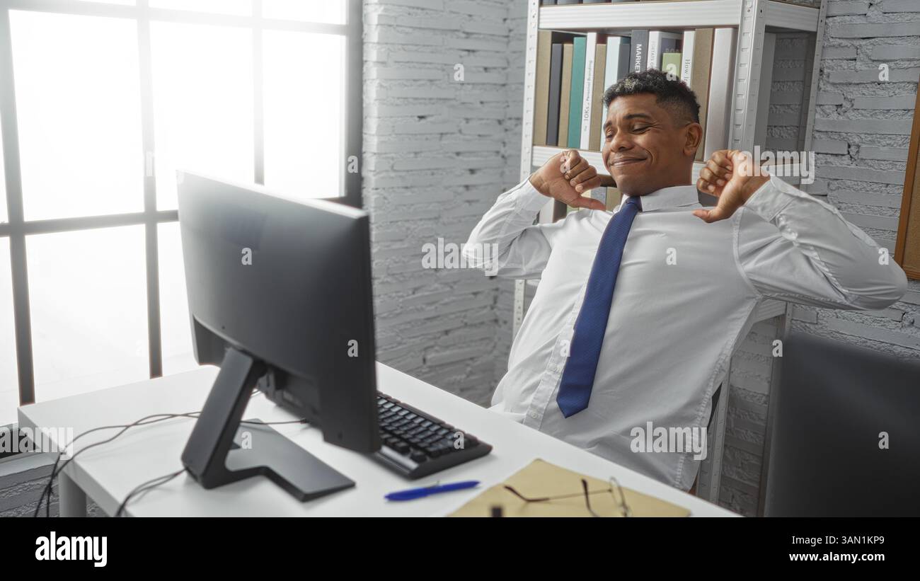 Young hispanic man stretches at desk in office room enjoying a break ...