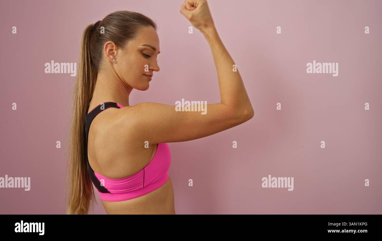 Woman flexing bicep in pink sports bra against solid pink wall, showcasing fitness and strength ...