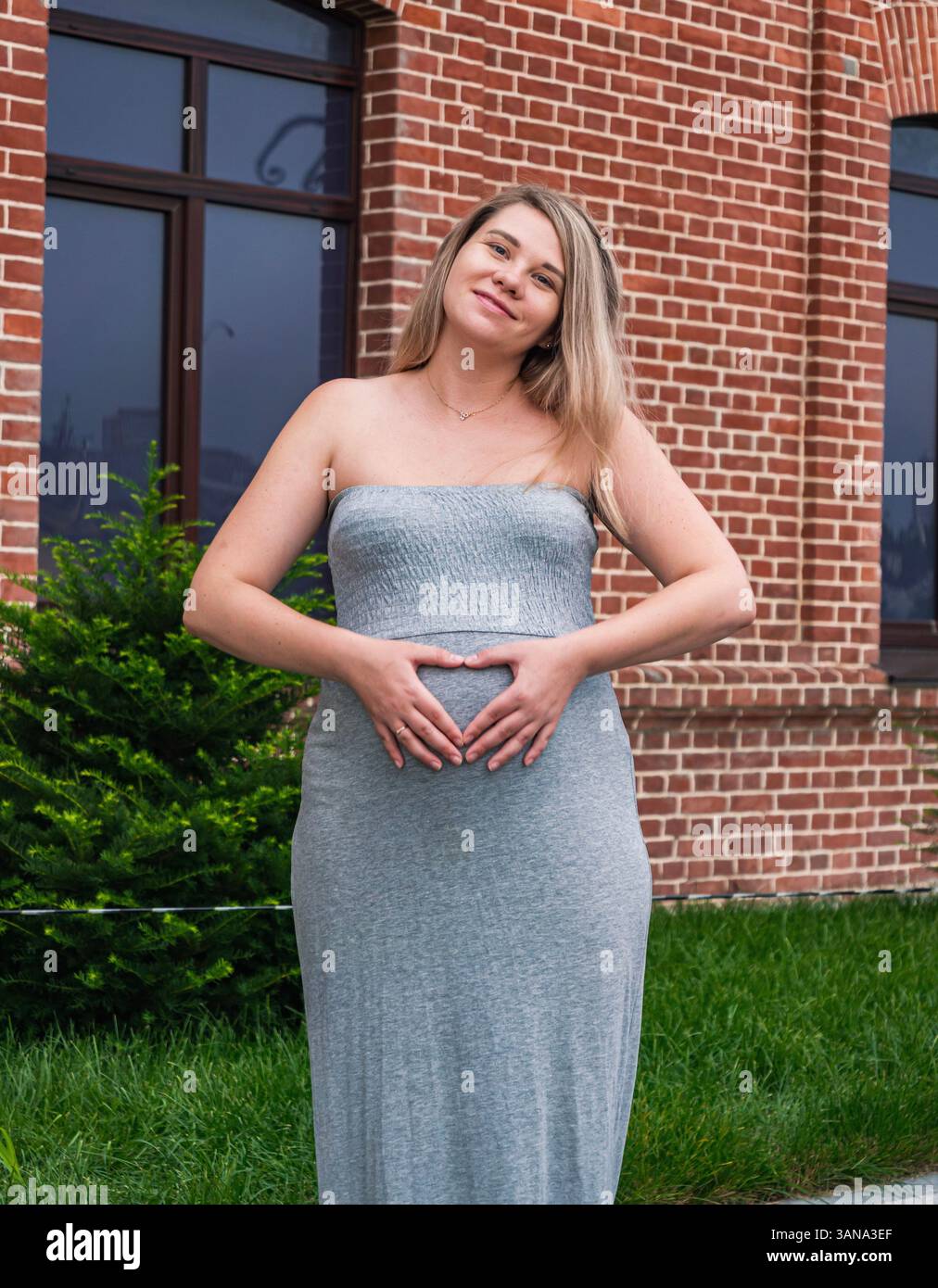 Pregnant woman cradles her baby bump while standing outdoors by a brick ...