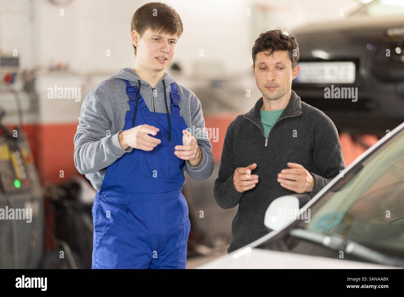 Young man car mechanic talks with man client in workshop and takes ...