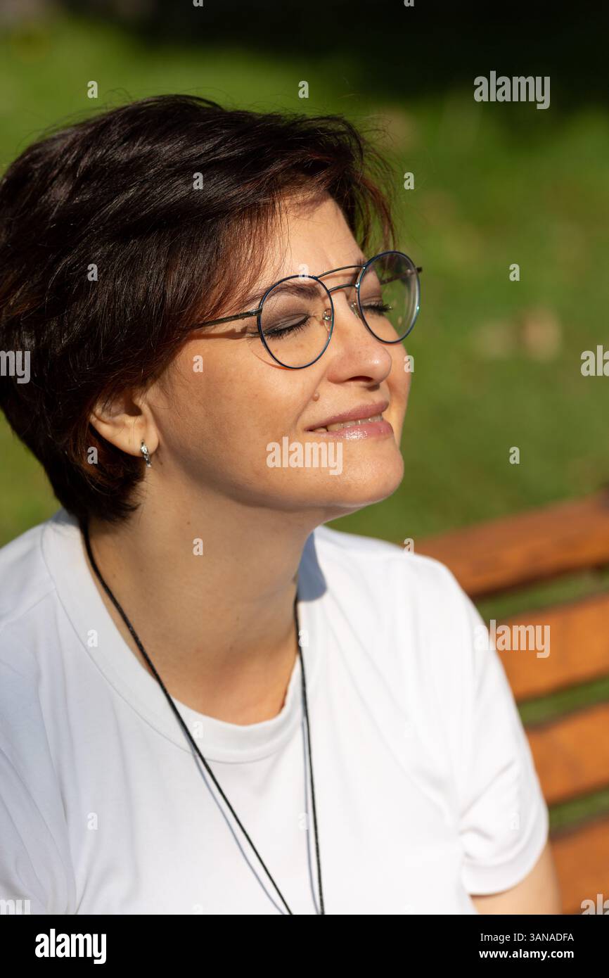 A beautiful middle-aged woman with glasses and short dark hair is sitting on a bench Stock Photo ...